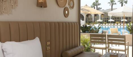 Quarto de hotel com cama, sofá, mesa e vista para uma piscina e palmeiras.
