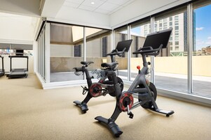 Uma academia com bicicletas ergométricas, esteira e amplas janelas com vista para a cidade.