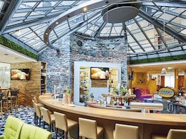 Uma área de bar com parede de pedra, teto alto com cobertura de vidro e um espaço de estar com cadeiras e um sofá.