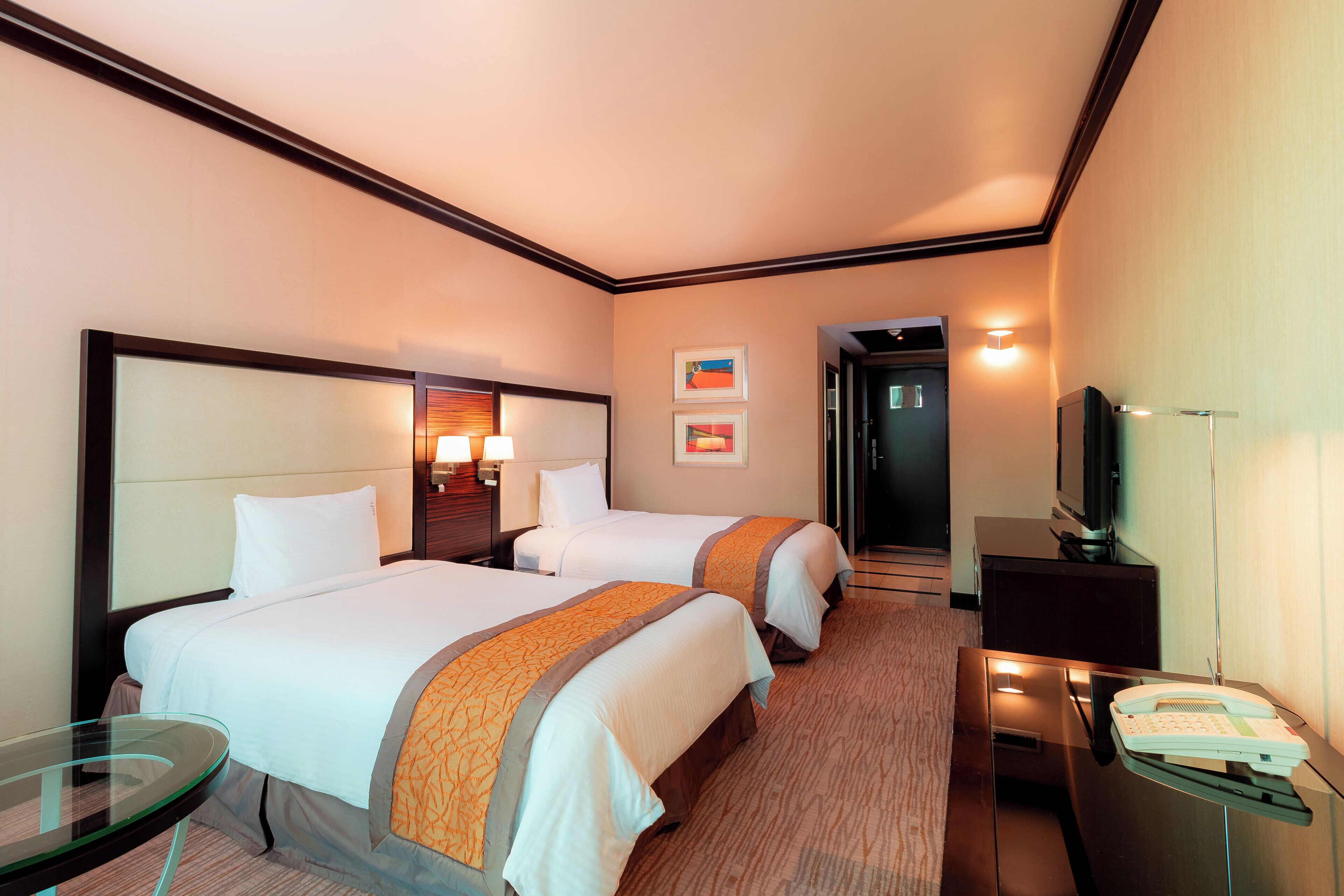Quarto de hotel com duas camas, uma mesa de vidro, uma escrivaninha com telefone e uma televisão.
