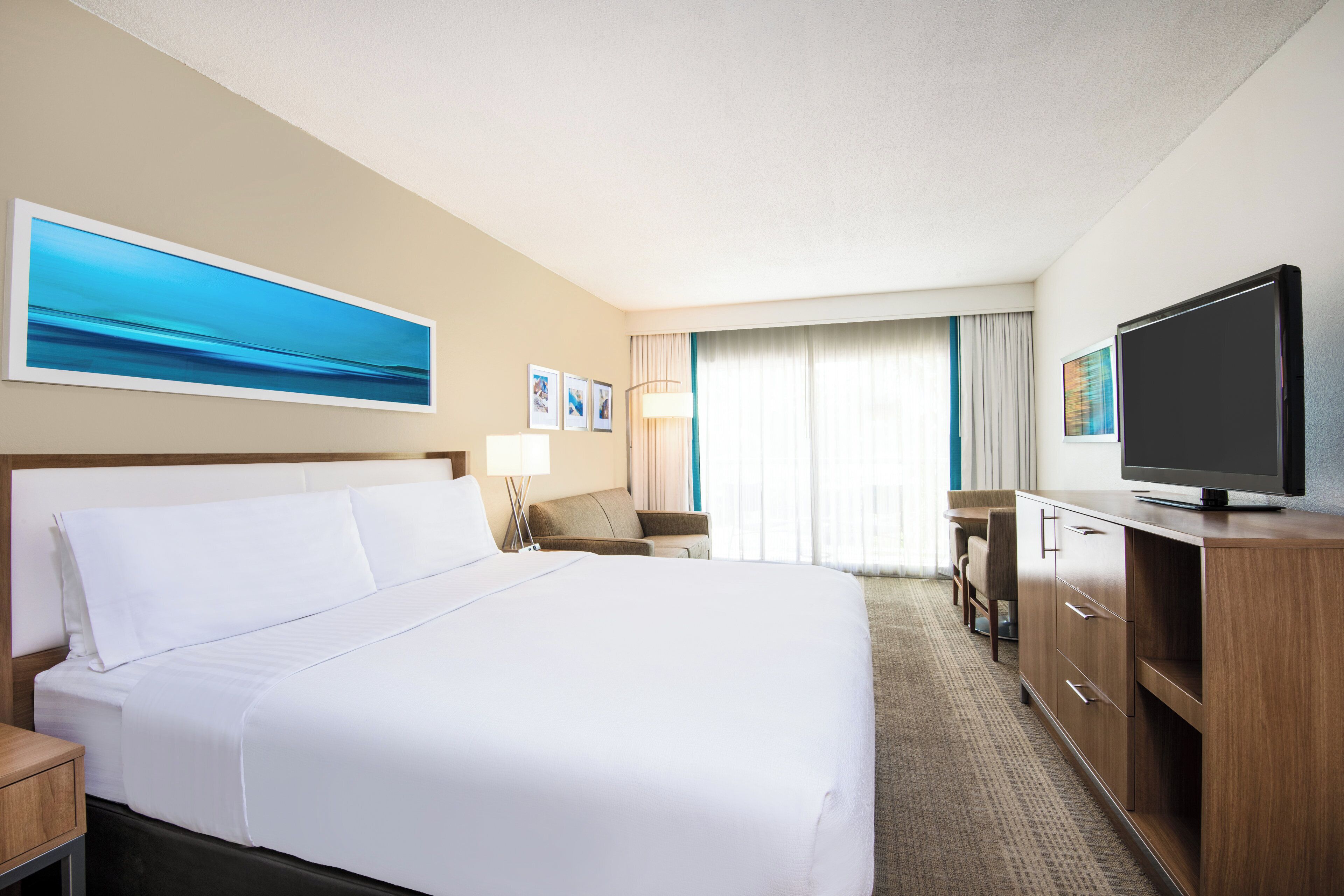 Quarto de hotel com uma cama grande, televisão, escrivaninha com cadeira e janela com cortinas.