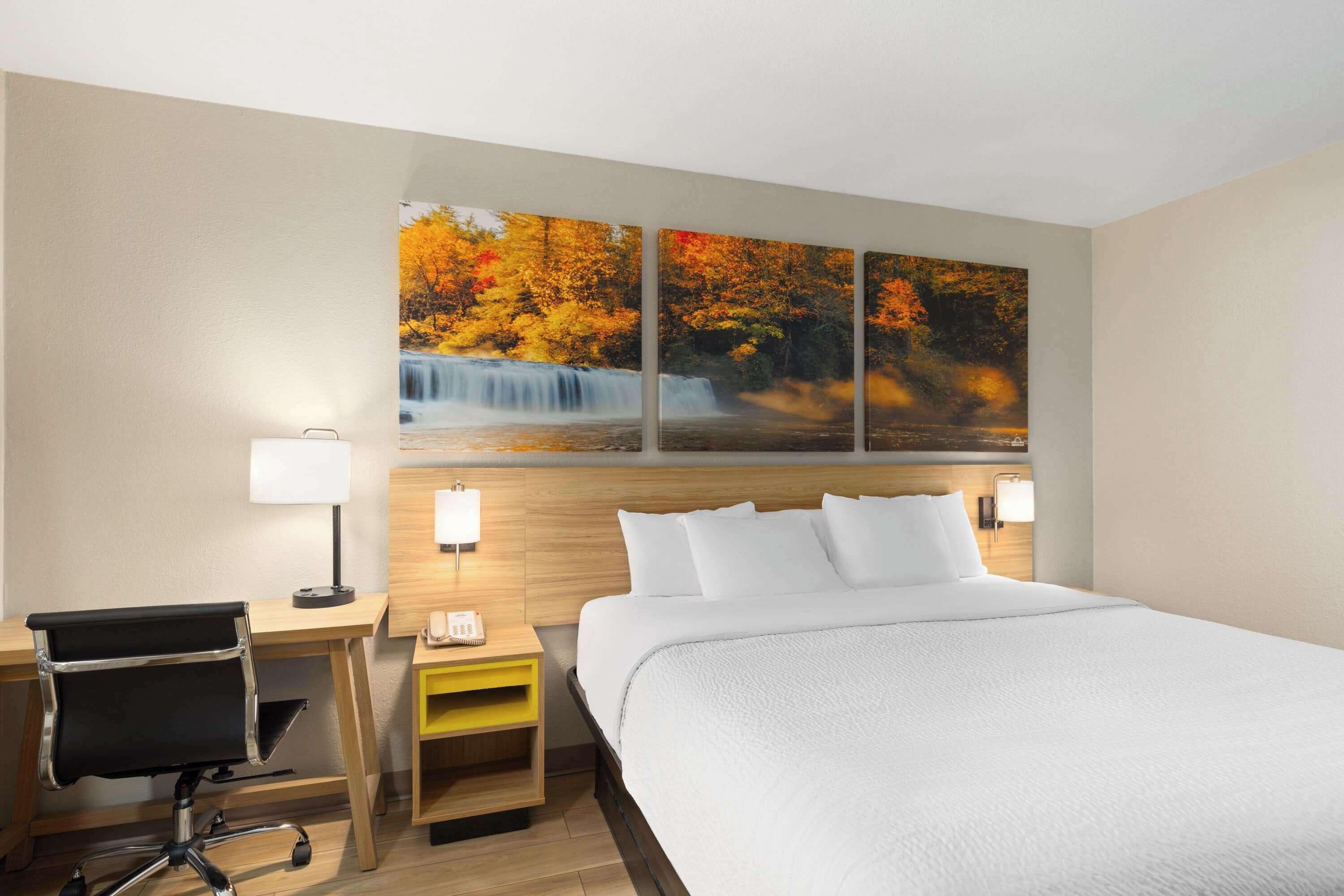 Quarto de hotel com uma cama grande, uma escrivaninha com cadeira, uma mesinha de cabeceira e uma luminária na parede.