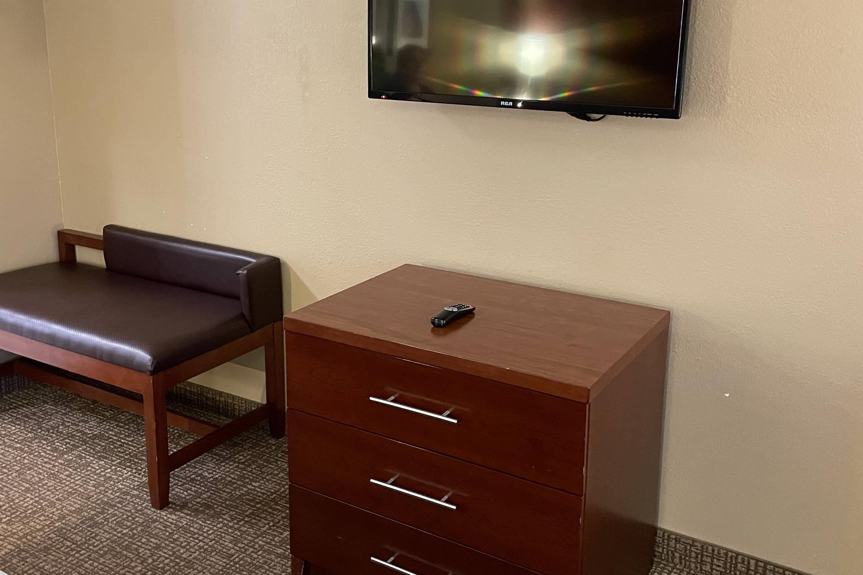Quarto de hotel com uma cama de solteiro, mesa de cabeceira de madeira, televisão fixada na parede e controle remoto na mesa.