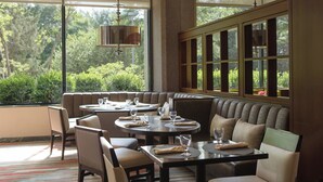 Restaurante com vista para o verde, com mesas redondas e cadeiras, um banco acolchoado e luminárias pendentes.