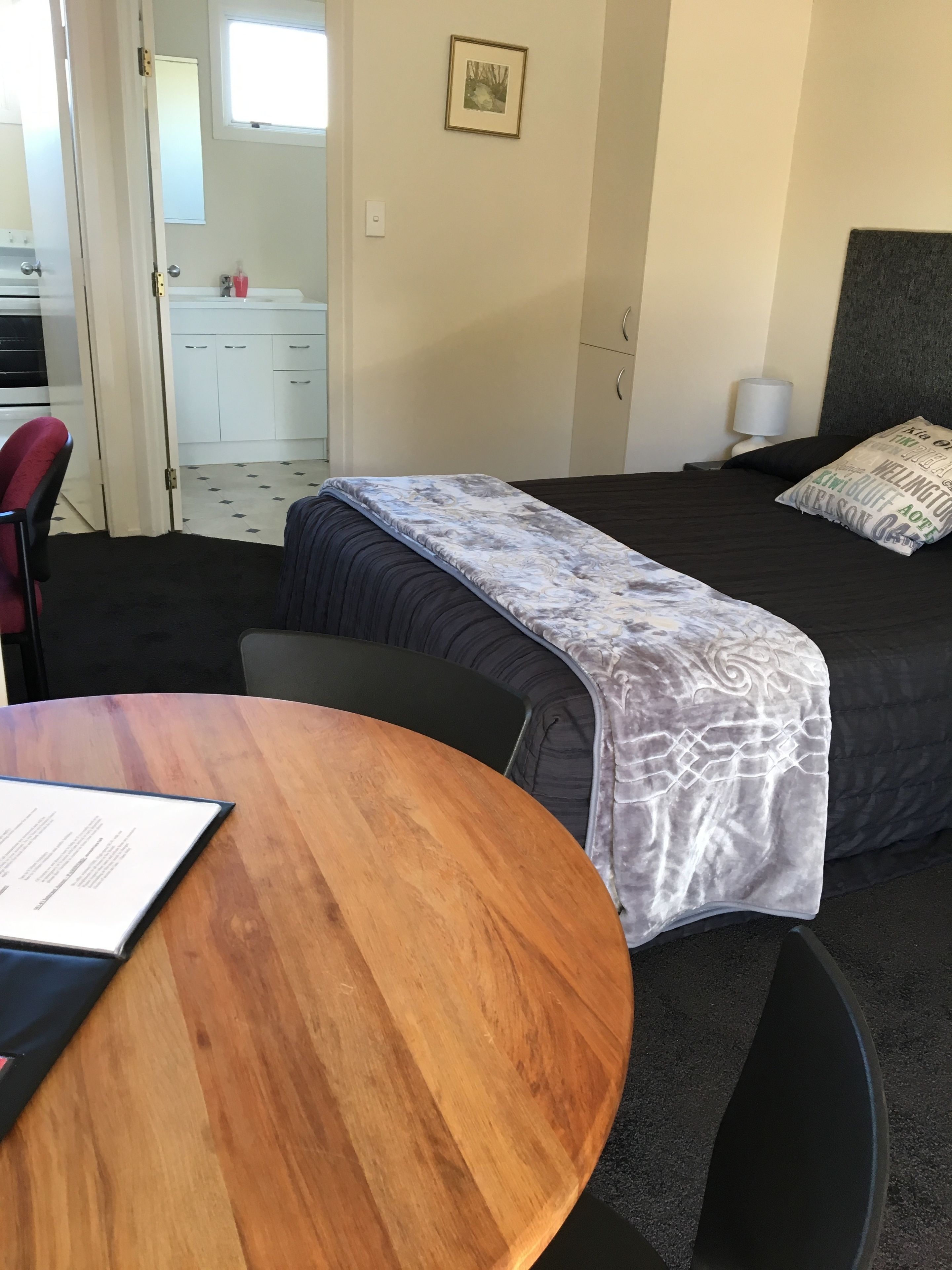 Quarto com uma cama, uma mesa de madeira redonda e uma kitchenette ao fundo.