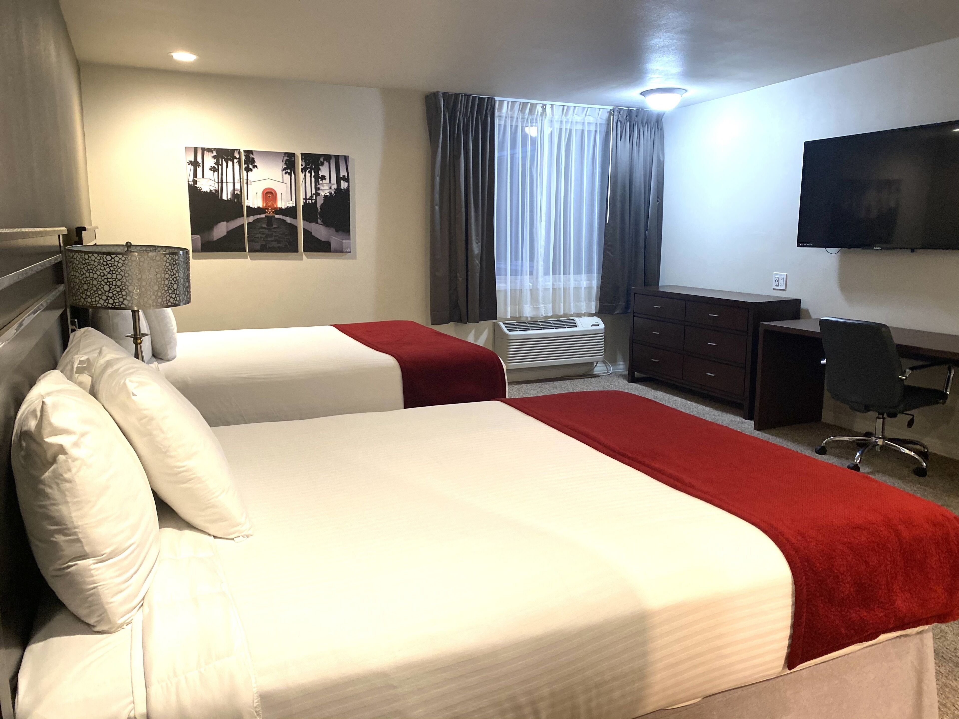 Quarto de hotel com duas camas, uma escrivaninha, uma cadeira, uma televisão e uma janela com cortinas.