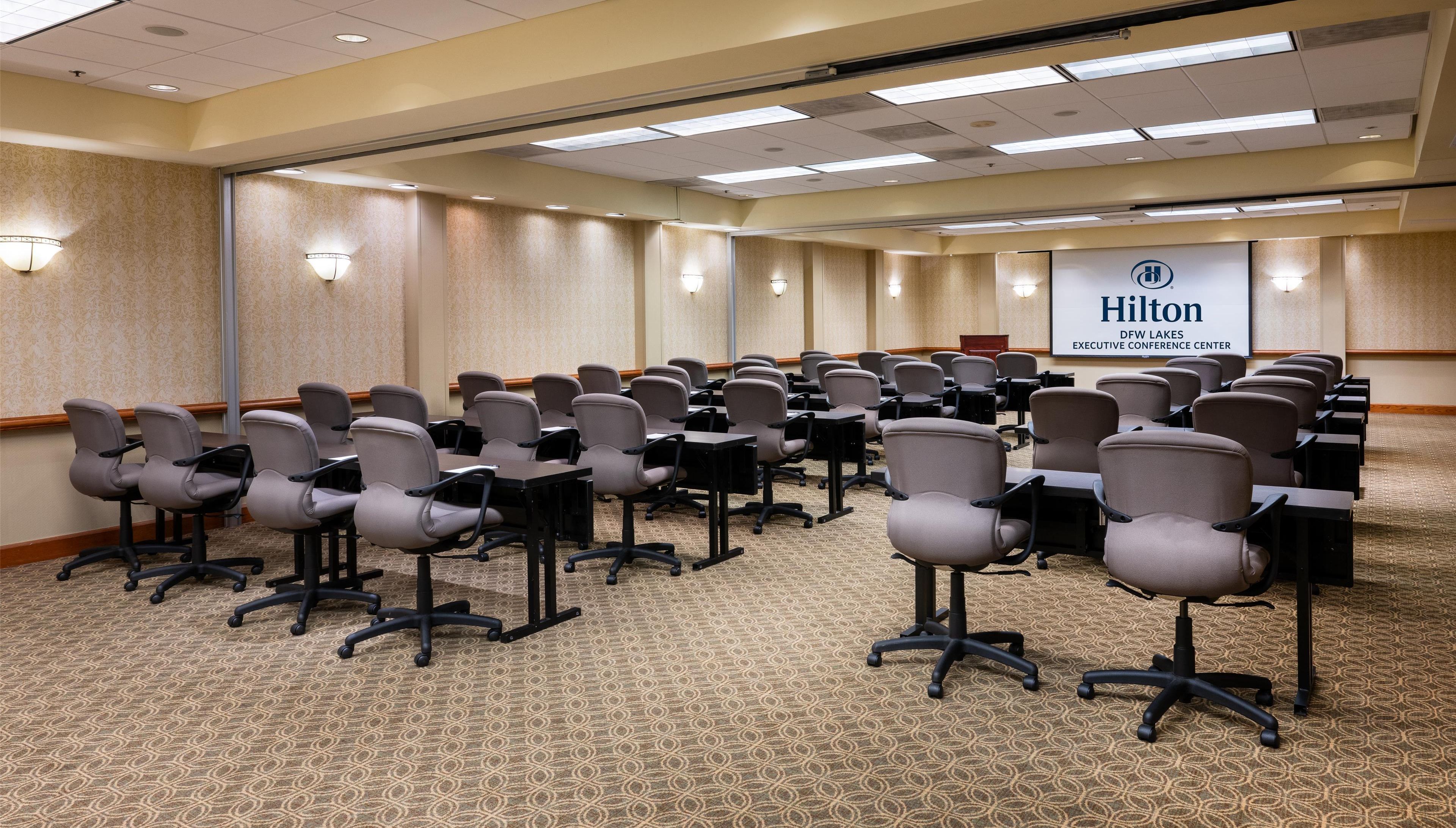 Uma sala de conferências com fileiras de cadeiras e mesas, piso coberto por carpete, e um banner com a inscrição "Hilton DFW Lakes Executive Conference Center".