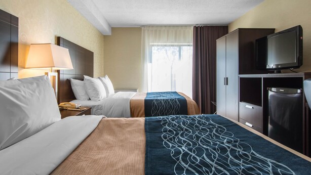 Quarto de hotel com duas camas, televisão, frigobar e uma janela grande com cortinas.