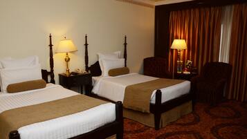 Quarto de hotel com duas camas, cada uma com estrutura de madeira e roupa de cama branca. Há duas mesas de cabeceira com luminárias, uma cadeira e uma janela grande com cortinas.