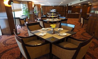 Uma sala de jantar de hotel com várias mesas, cadeiras, uma tela de televisão e uma planta.