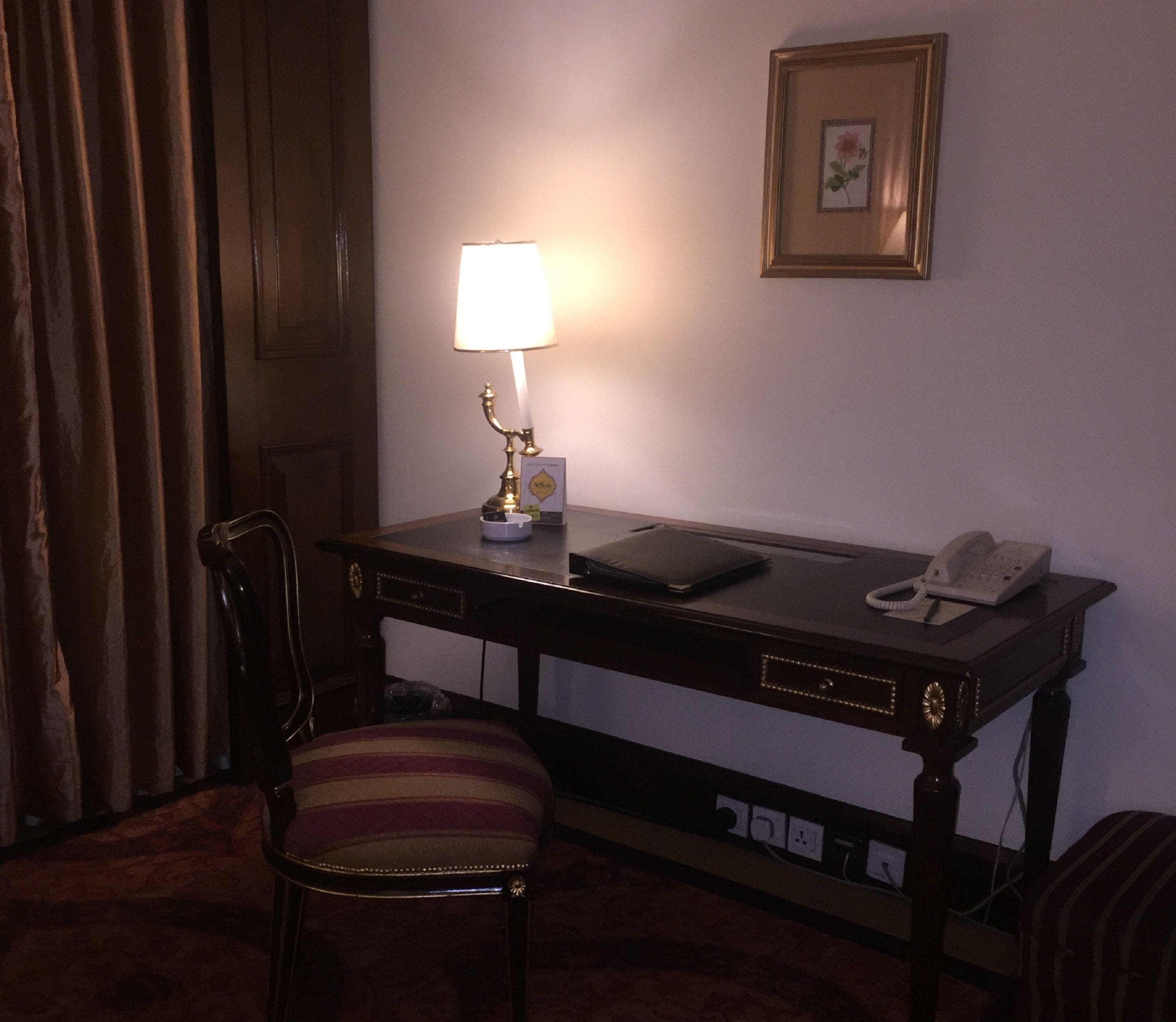 Quarto de hotel com uma escrivaninha de madeira, luminária, telefone e uma cadeira.
