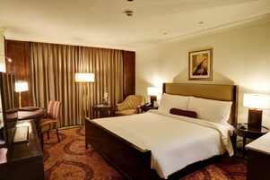 Quarto de hotel com uma cama grande, uma cadeira, uma escrivaninha e um abajur.
