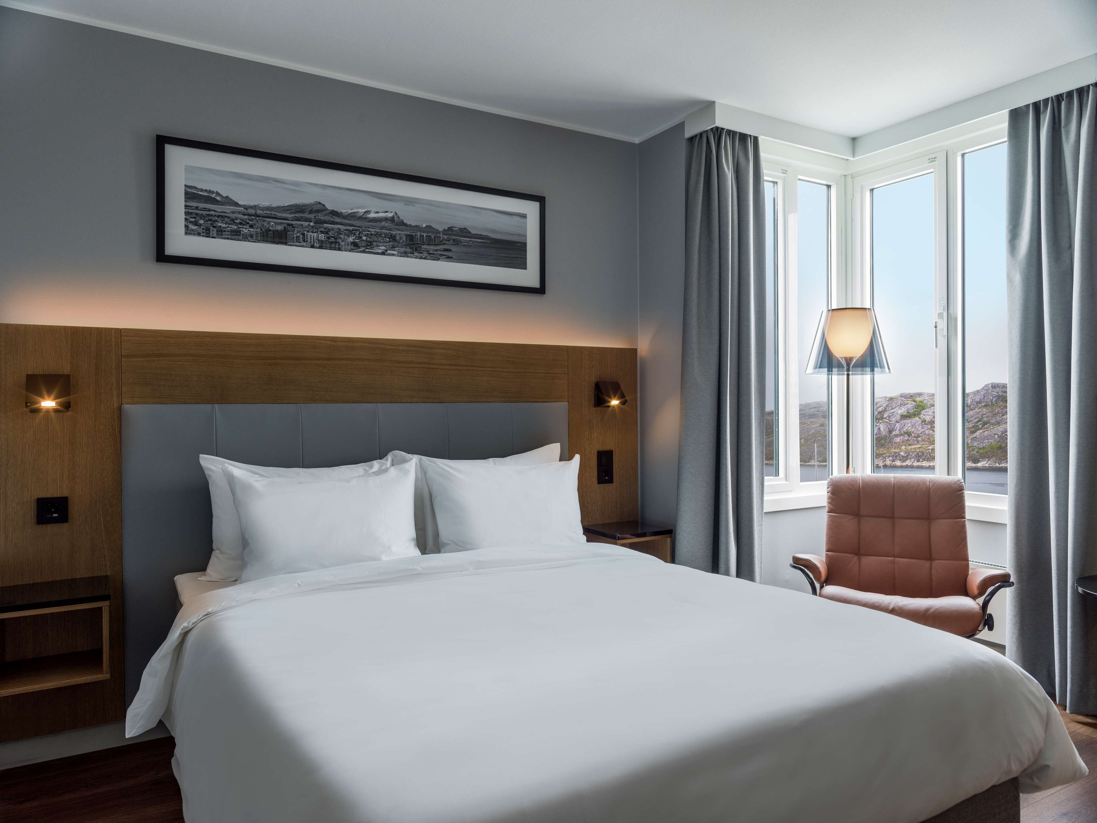 Quarto de hotel com uma cama grande, cabeceira, quadro com vista da cidade, janela com cortinas e uma cadeira.