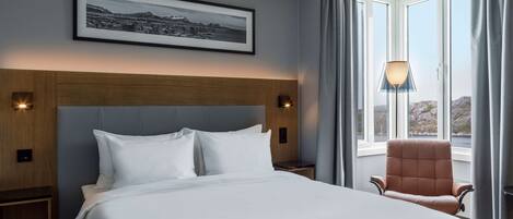 Quarto de hotel com uma cama grande, cabeceira, quadro com vista da cidade, janela com cortinas e uma cadeira.