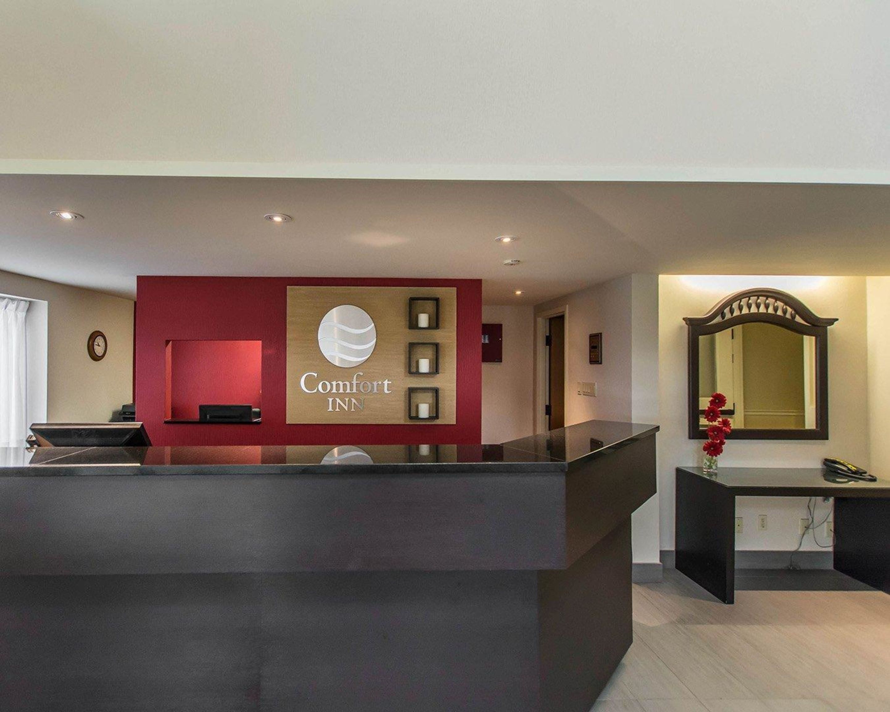 Uma área de recepção de hotel com uma parede de destaque vermelha, a placa Comfort Inn, um balcão de recepção, um espelho e um vaso com flores.