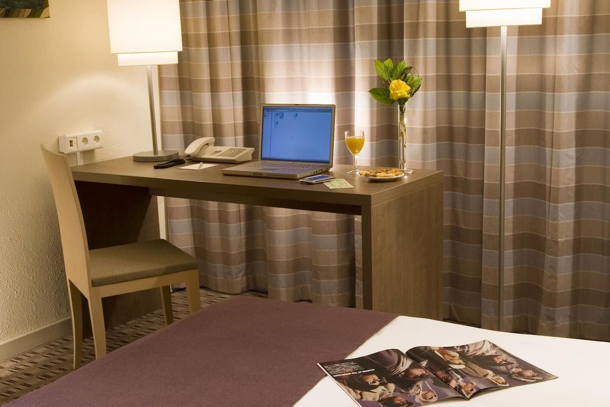 Quarto de hotel com cama, escrivaninha com laptop, telefone, vaso com flores e revista na cama.