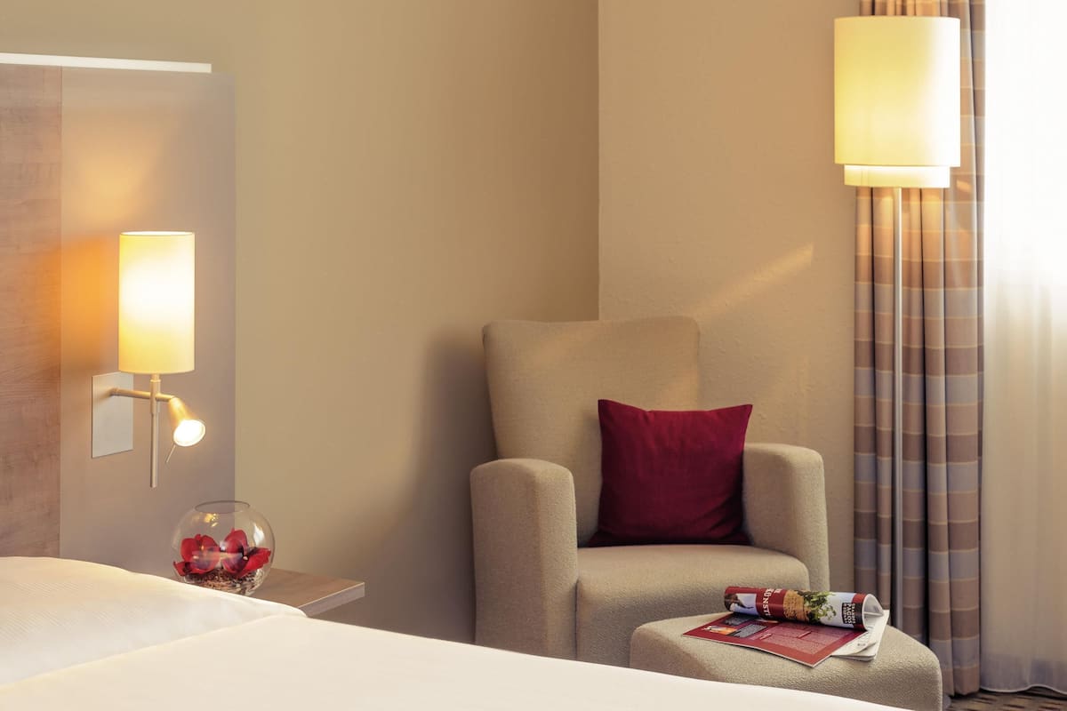 Quarto de hotel com uma cama, uma poltrona com almofada vermelha, mesa de cabeceira com abajur e um copo de água.