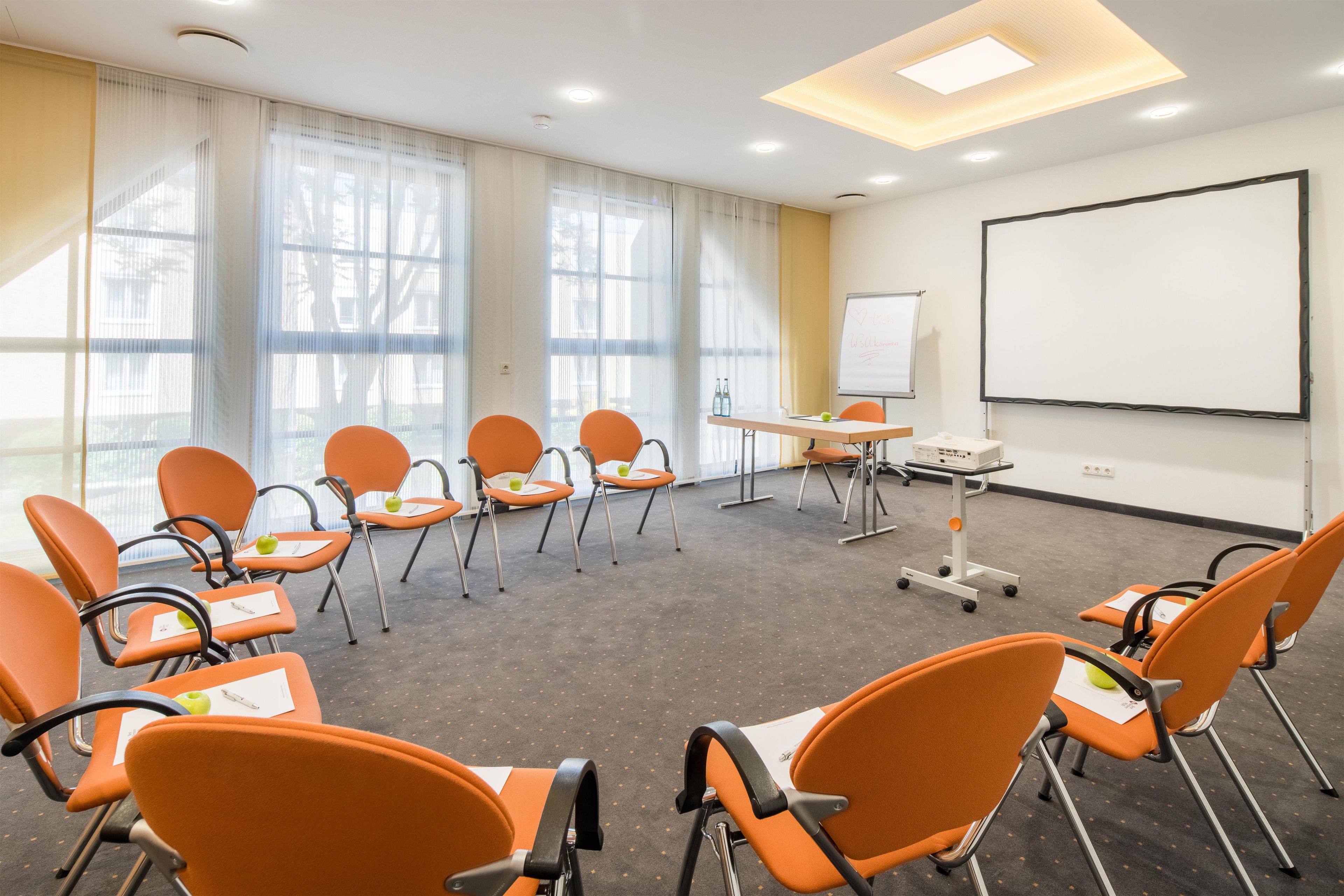 Sala de conferência com lousa, projetor e várias cadeiras laranja com documentos.