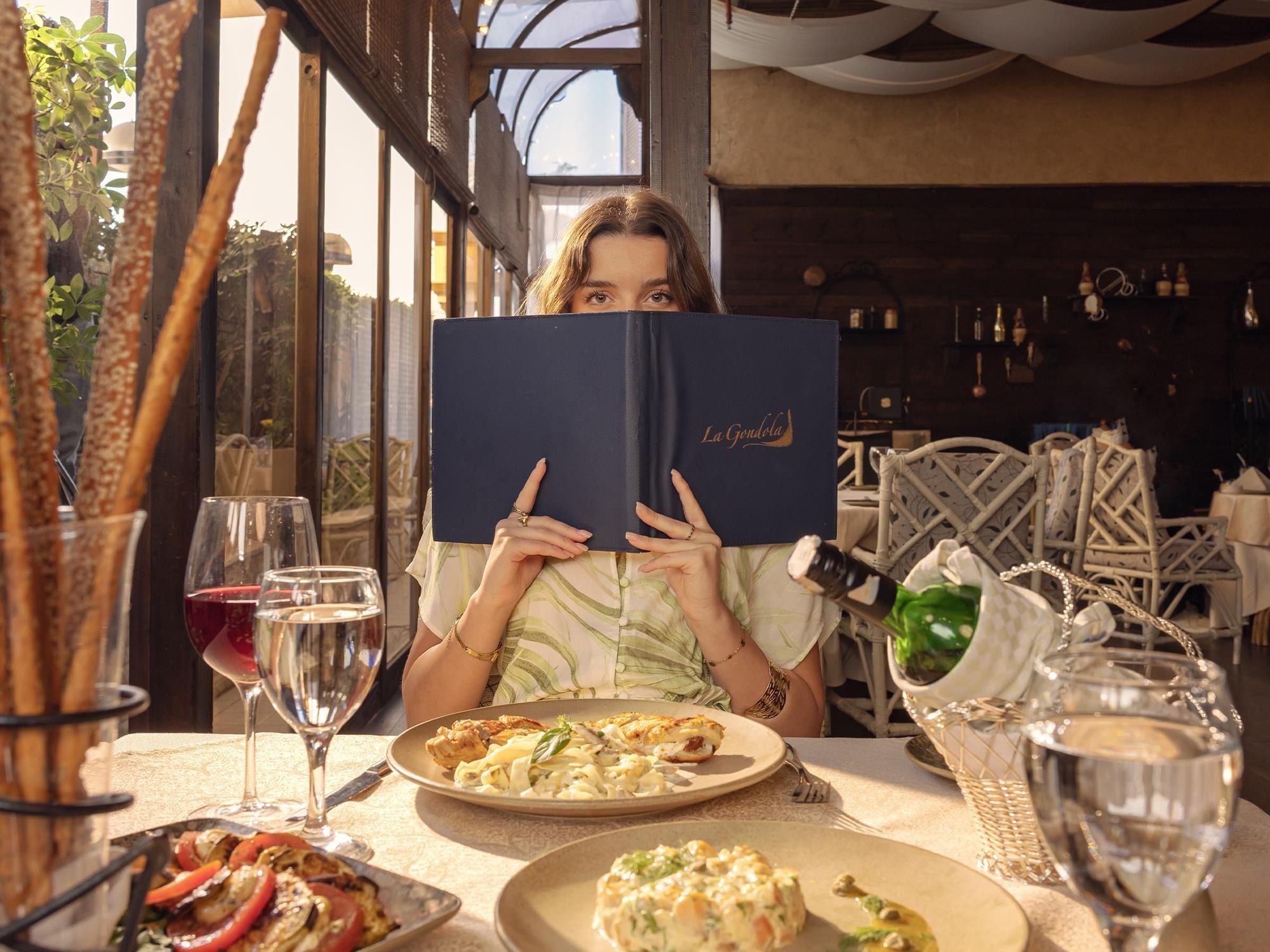 Uma pessoa sentada à mesa com um menu, copos de vinho e pratos de comida.