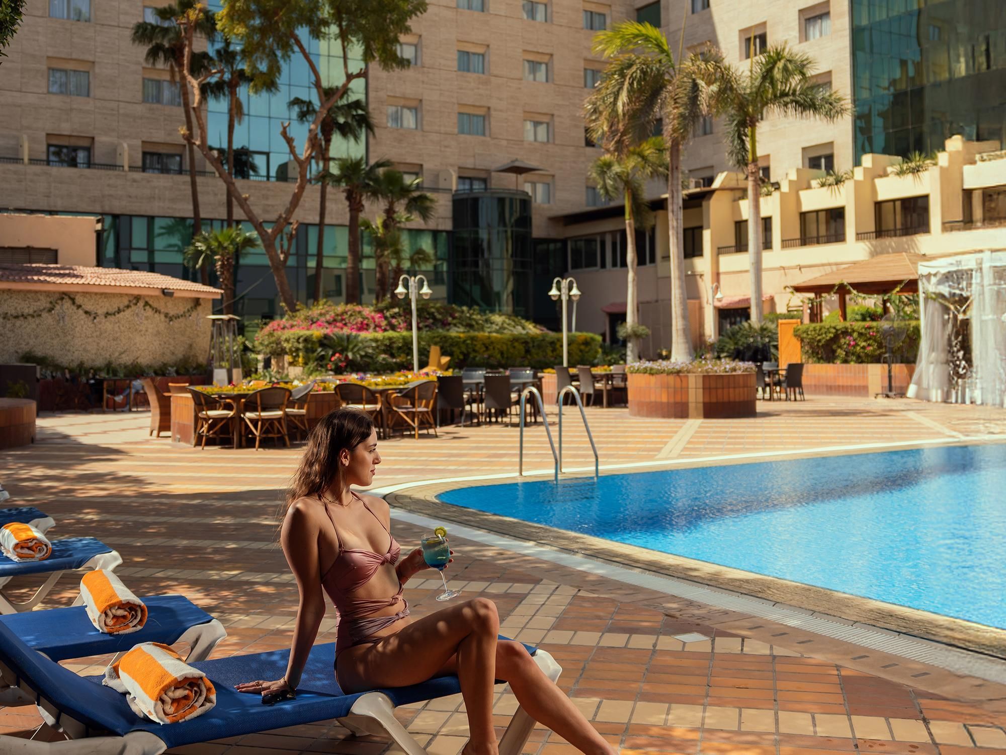 Uma área à beira da piscina com espreguiçadeiras, uma piscina e um edifício com vários andares ao fundo.