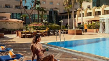 Uma área à beira da piscina com espreguiçadeiras, uma piscina e um edifício com vários andares ao fundo.