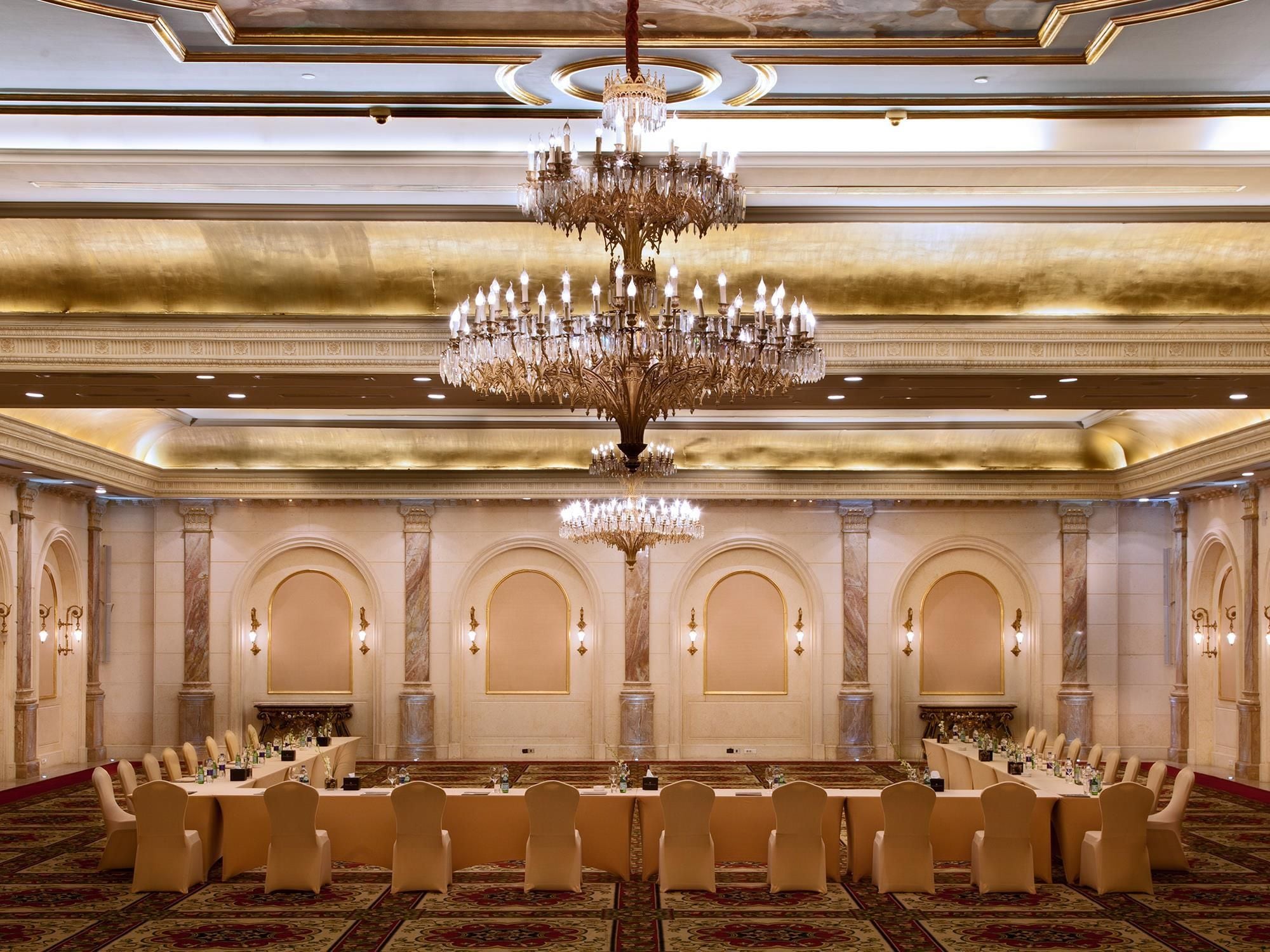 Uma imponente sala de banquetes com um grande candelabro, colunas ricamente decoradas e uma longa mesa posta para um evento formal.
