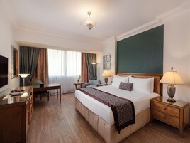 Quarto de hotel com cama, mesas de cabeceira, escrivaninha, cadeira, televisão e uma grande janela com cortinas.