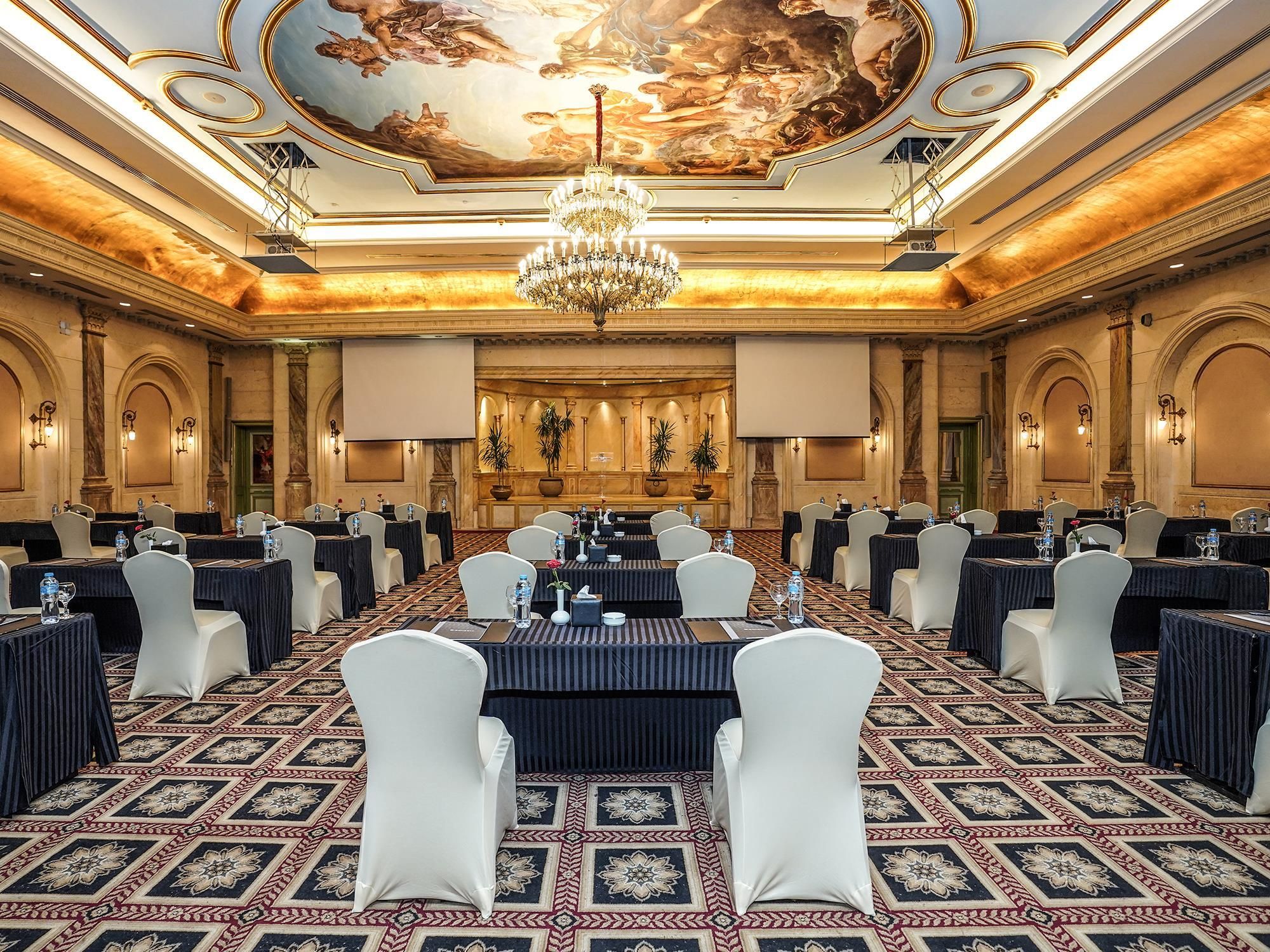 Uma sala de conferências ampla com teto alto, design ornamental no teto e um candelabro. Mesas dispostas com cadeiras, garrafas de água e placas de identificação.