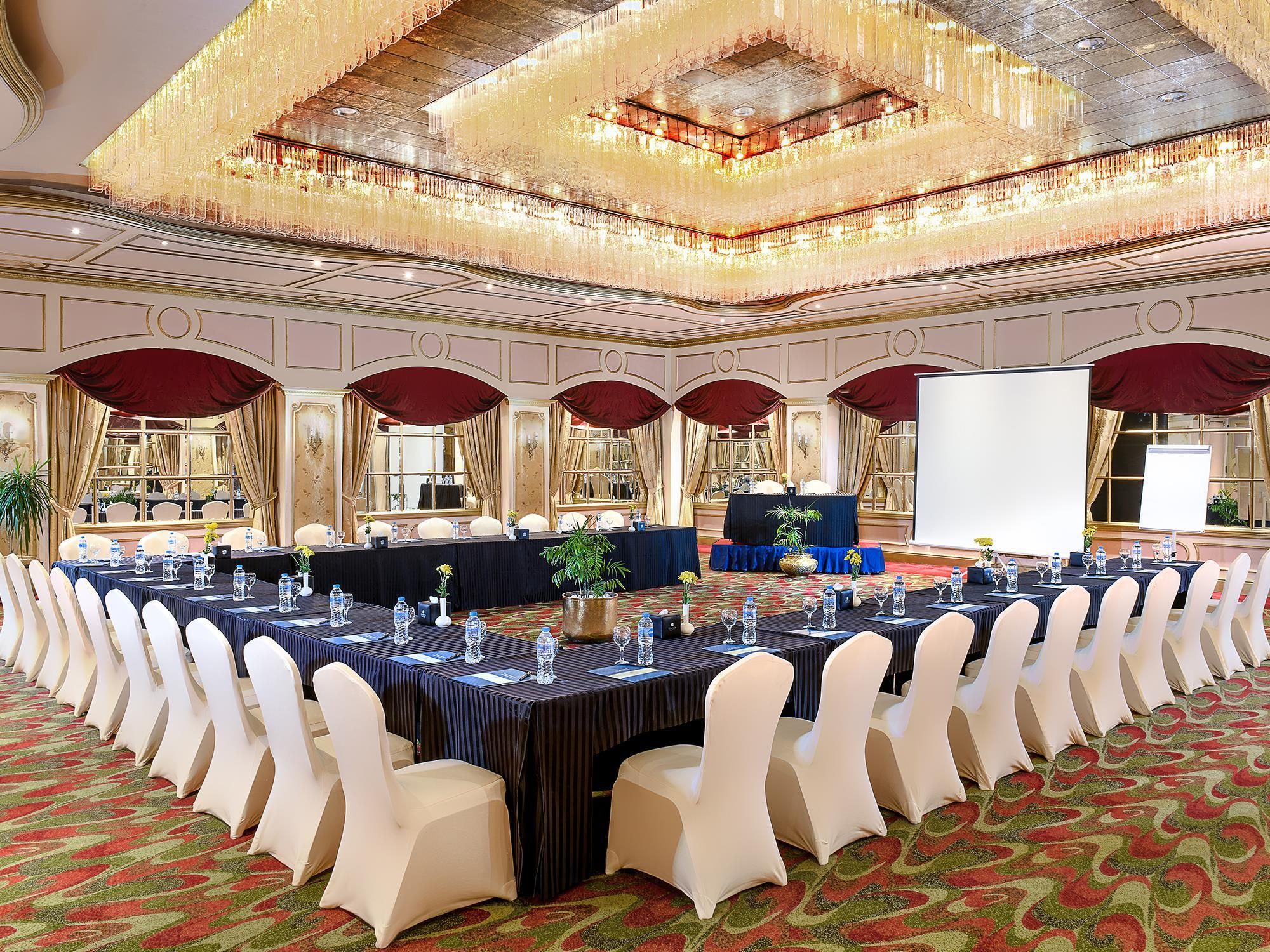 Uma sala de banquetes com mesas longas, cadeiras brancas e um palco para apresentações.