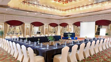 Uma sala de banquetes com mesas longas, cadeiras brancas e um palco para apresentações.