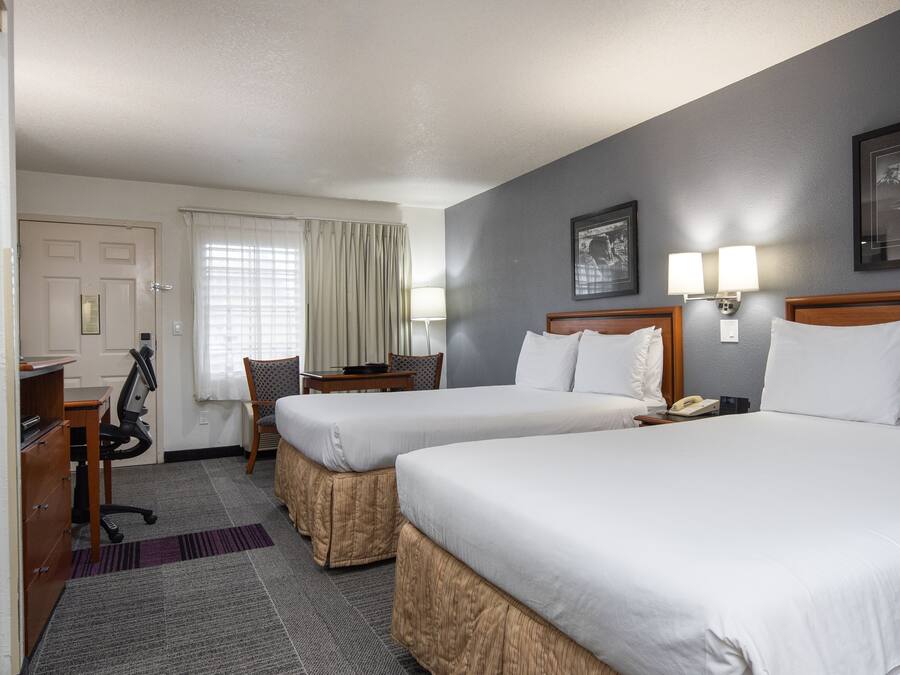 Quarto de hotel com duas camas, uma escrivaninha, uma cadeira e uma luminária de mesa.