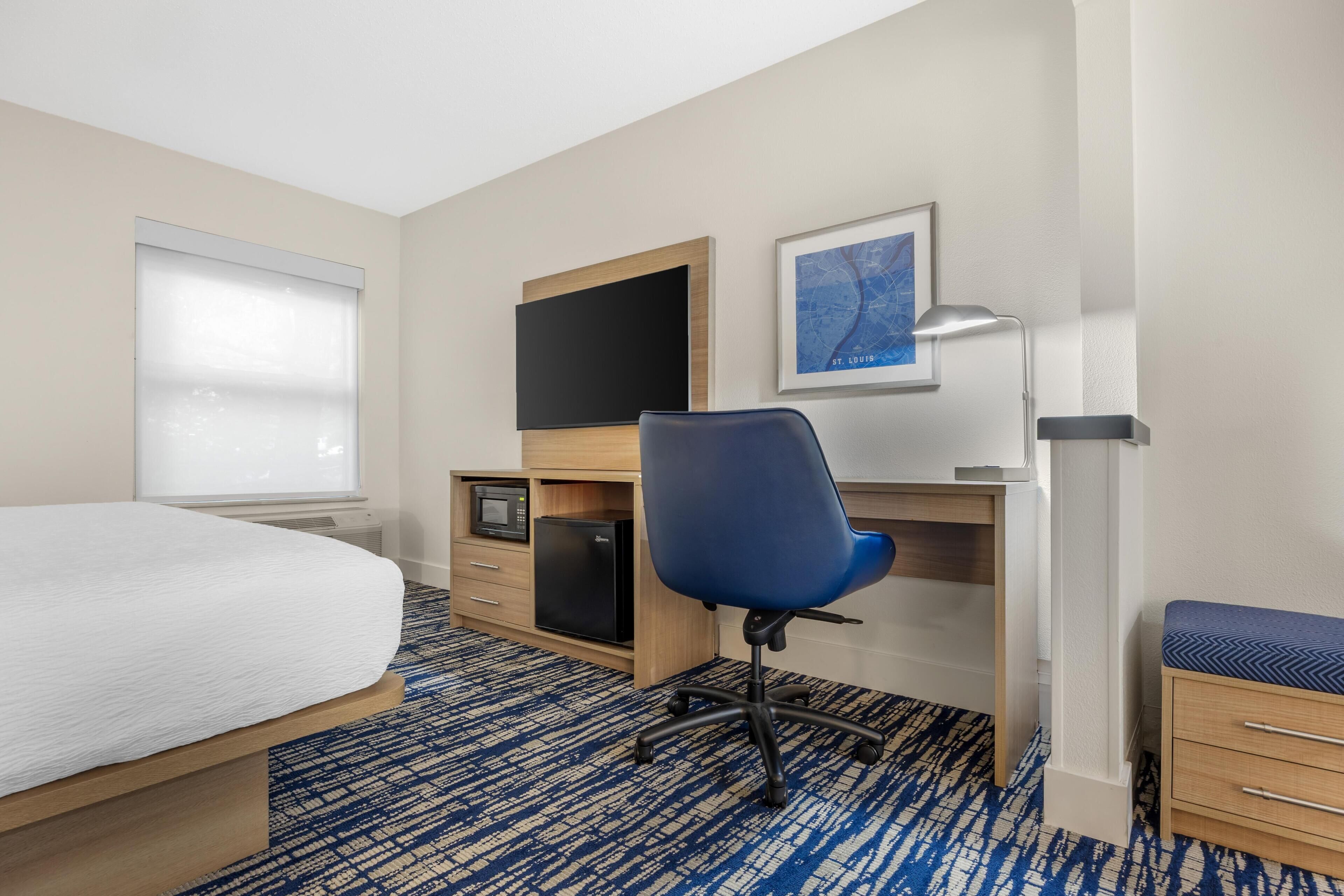 Quarto de hotel com cama, escrivaninha, cadeira, micro-ondas e um mapa emoldurado na parede.