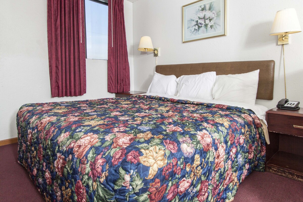 Quarto de hotel com colcha estampada de flores, duas luminárias de cabeceira, uma obra de arte floral emoldurada na parede e um telefone no criado-mudo.