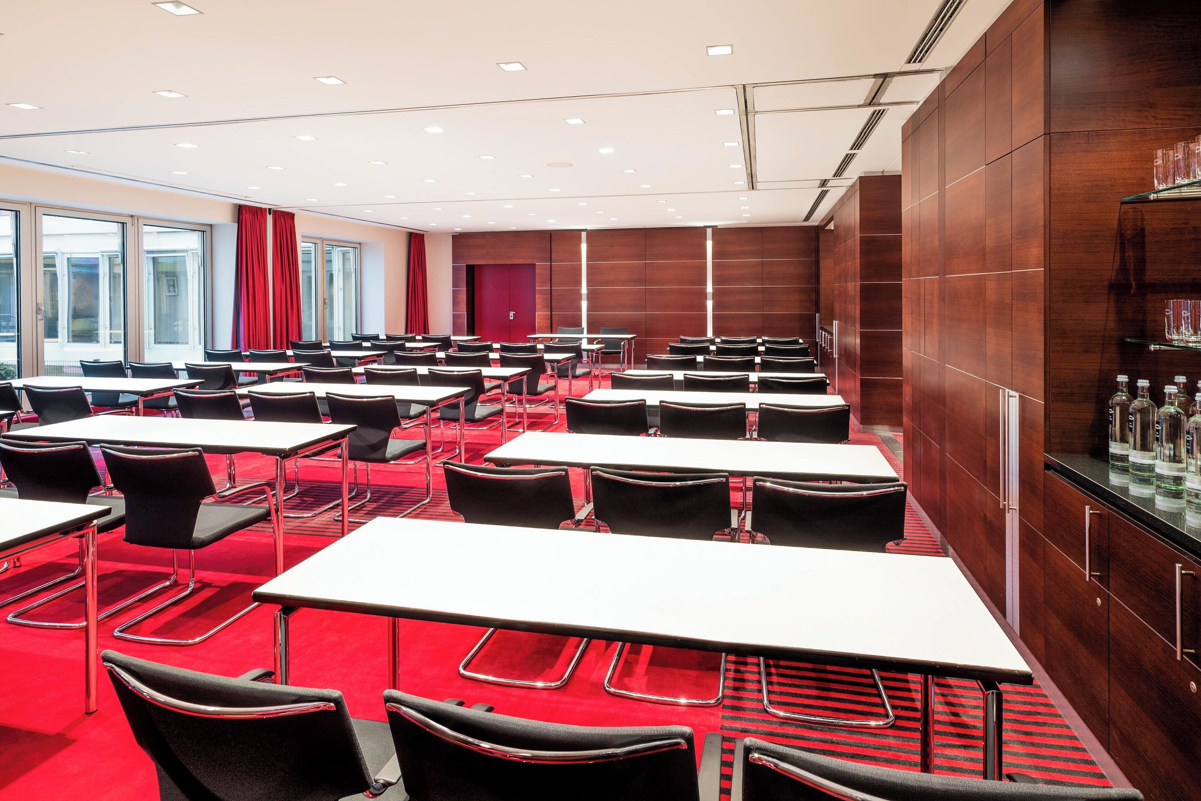 Uma sala de conferências com mesas e cadeiras, janelas amplas e um tapete vermelho.