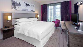 Quarto de hotel com uma cama grande, uma escrivaninha com cadeira, uma televisão e um telefone na mesinha de cabeceira.