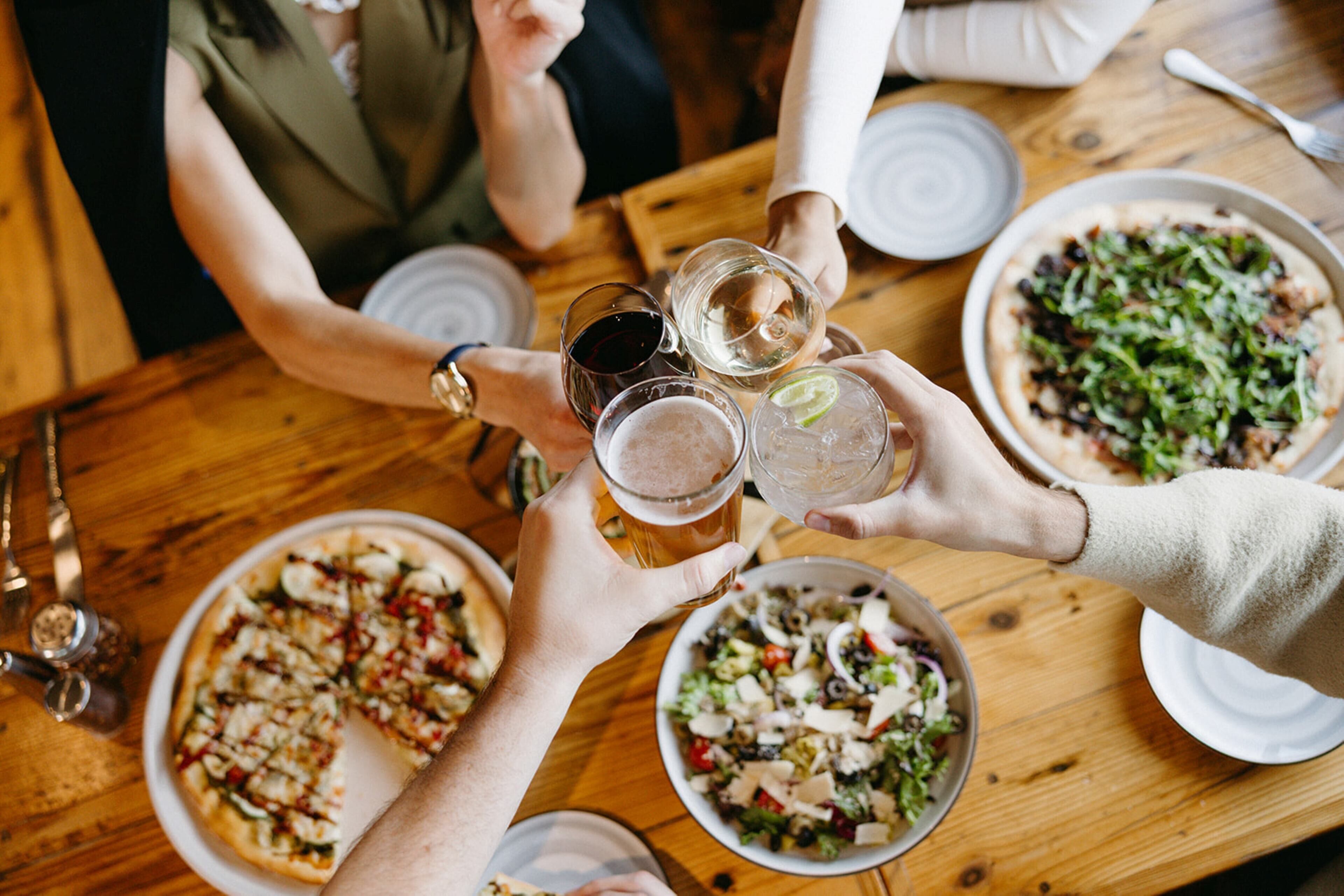 Um grupo de pessoas brindando com bebidas em volta de uma mesa de madeira com pizza e salada.