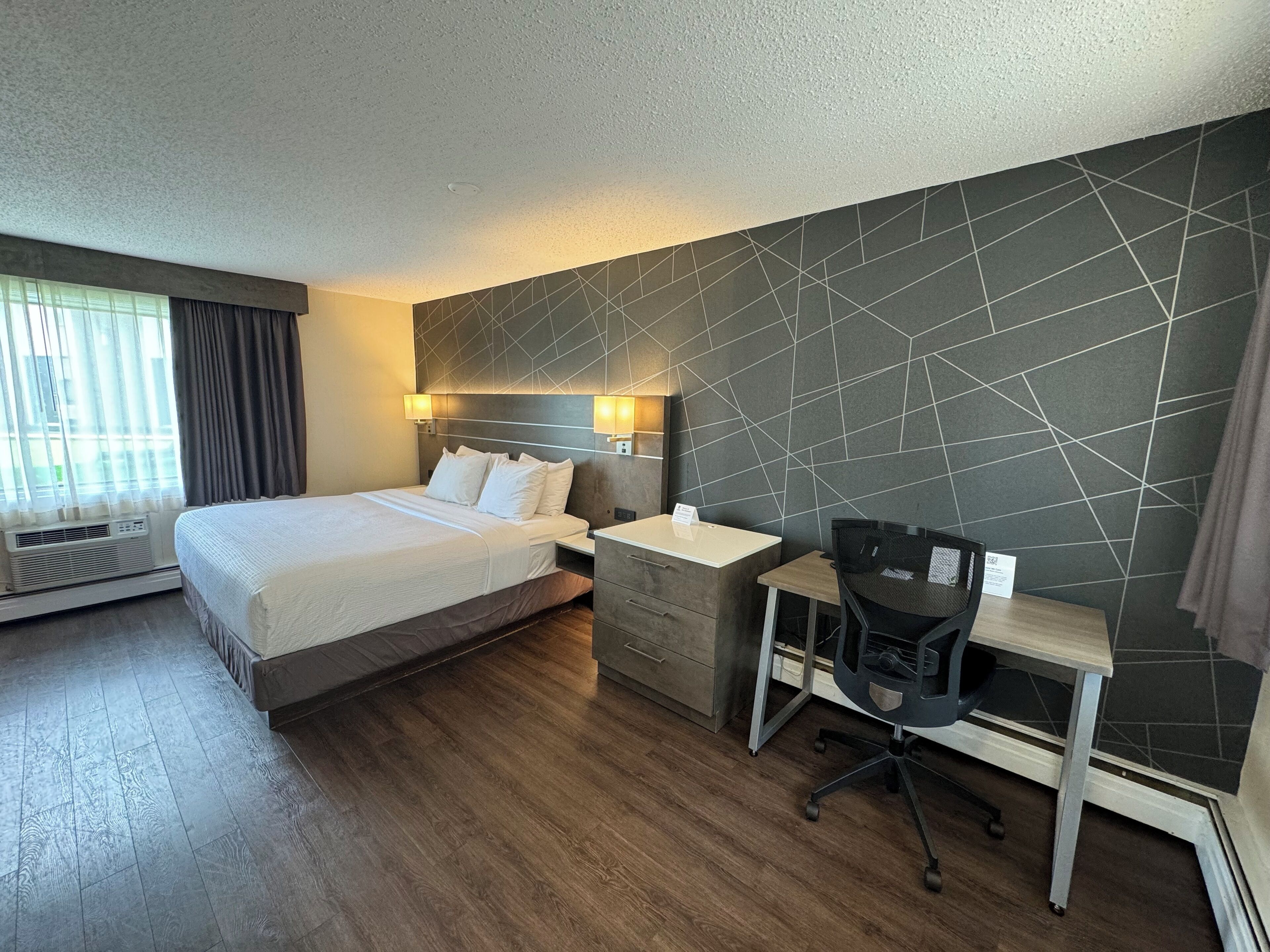 Quarto de hotel com cama, mesa de cabeceira, escrivaninha, cadeira e parede com padrão geométrico.