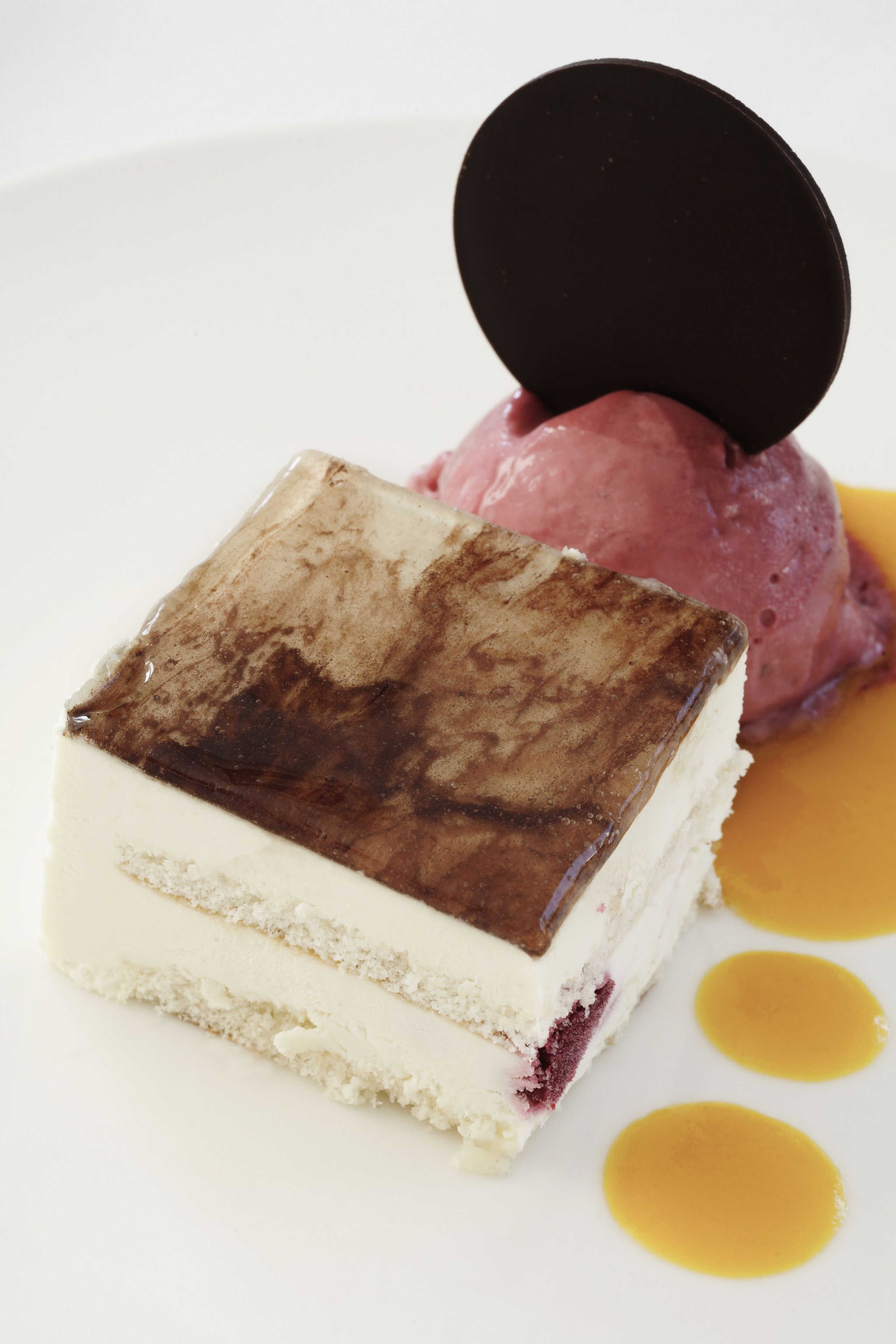 Sobremesa com camada de chocolate, creme branco e uma bola de sorvete rosa em um prato branco.