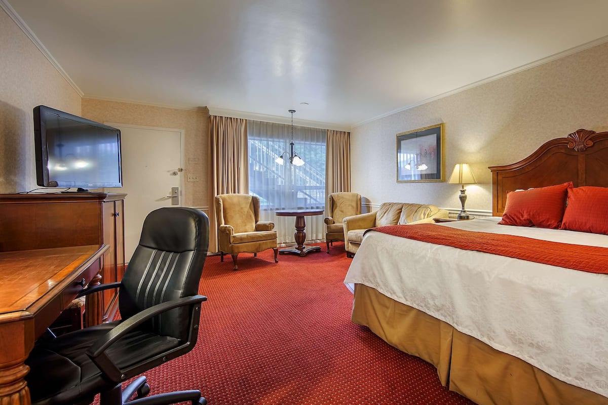Quarto de hotel com uma cama grande, uma mesa com televisão, duas poltronas, uma pequena mesa e uma janela com cortinas.