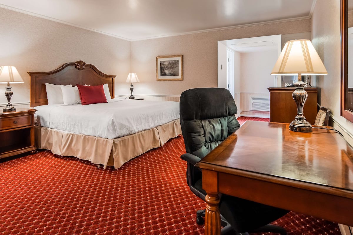 Quarto de hotel com uma cama grande, uma escrivaninha, duas mesas de cabeceira com luminárias, uma cadeira e um chão coberto por um tapete vermelho.