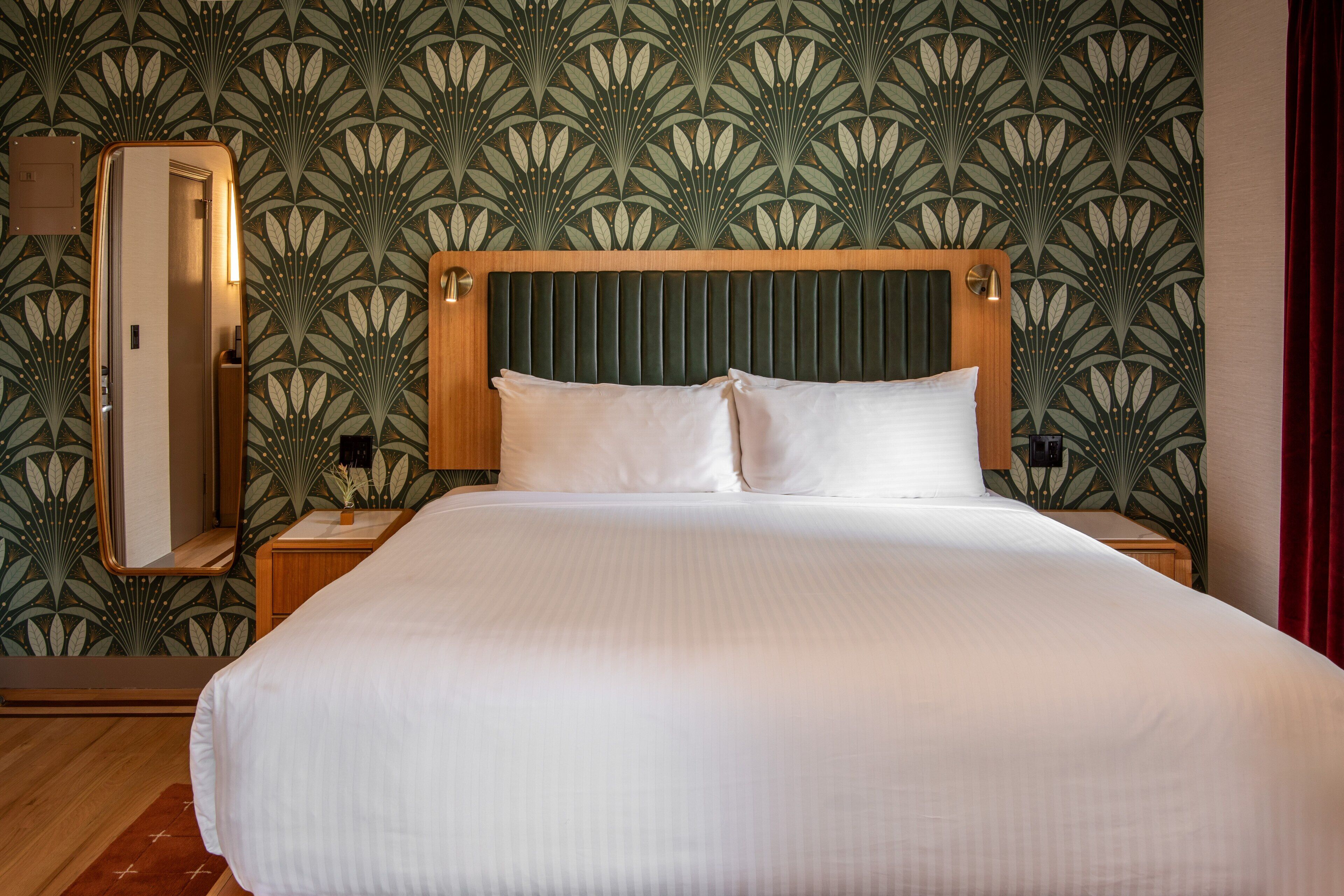 Quarto de hotel com uma cama grande, cabeceira de madeira, mesa de cabeceira, espelho e papel de parede estampado.