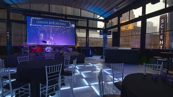Um salão amplo com palco, cadeiras e mesas, exibindo uma tela grande com a inscrição 'Edison Ballroom'.