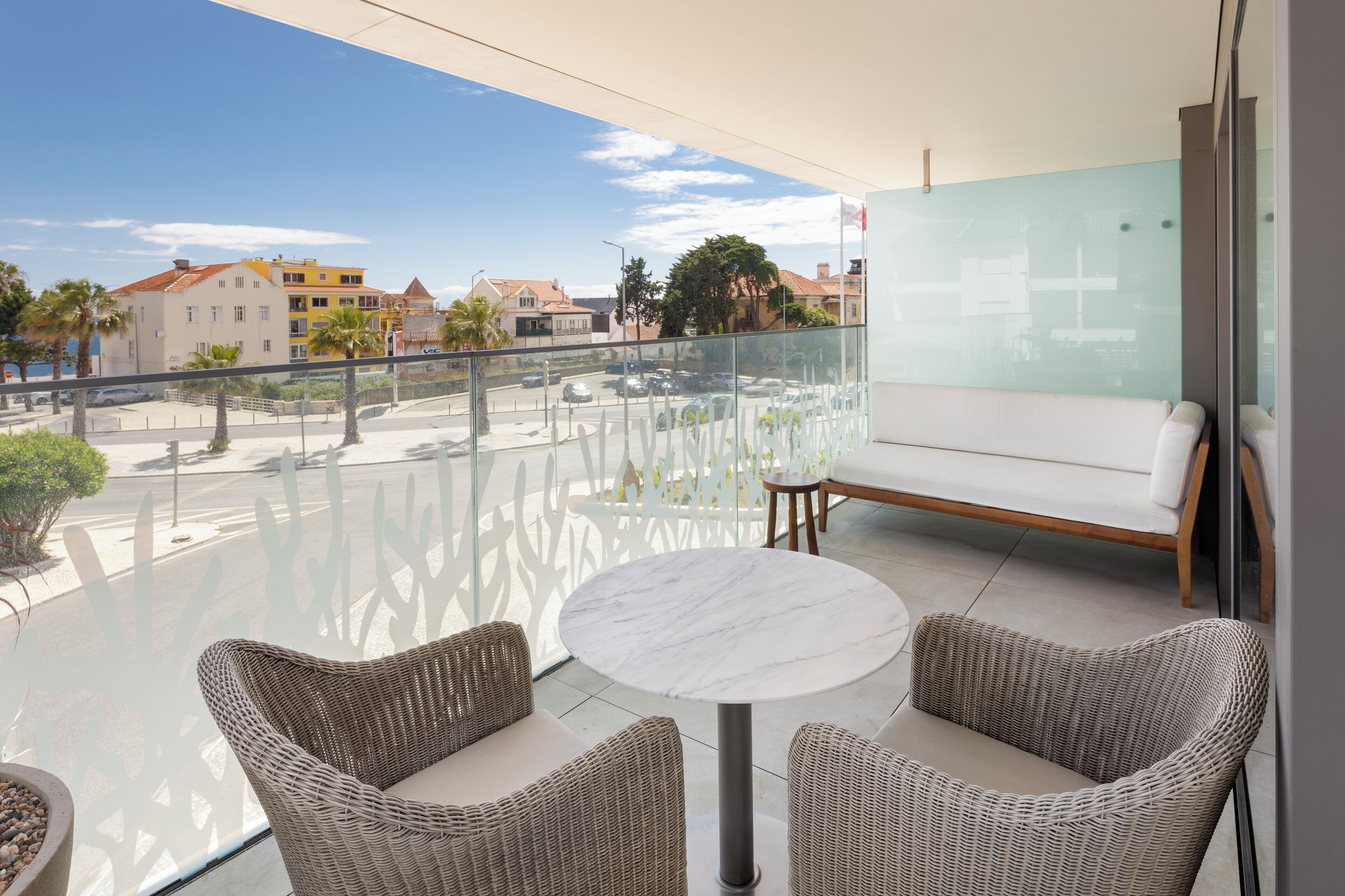 Uma varanda com mobiliário de vime, uma mesa de mármore e vista para a rua e edifícios.