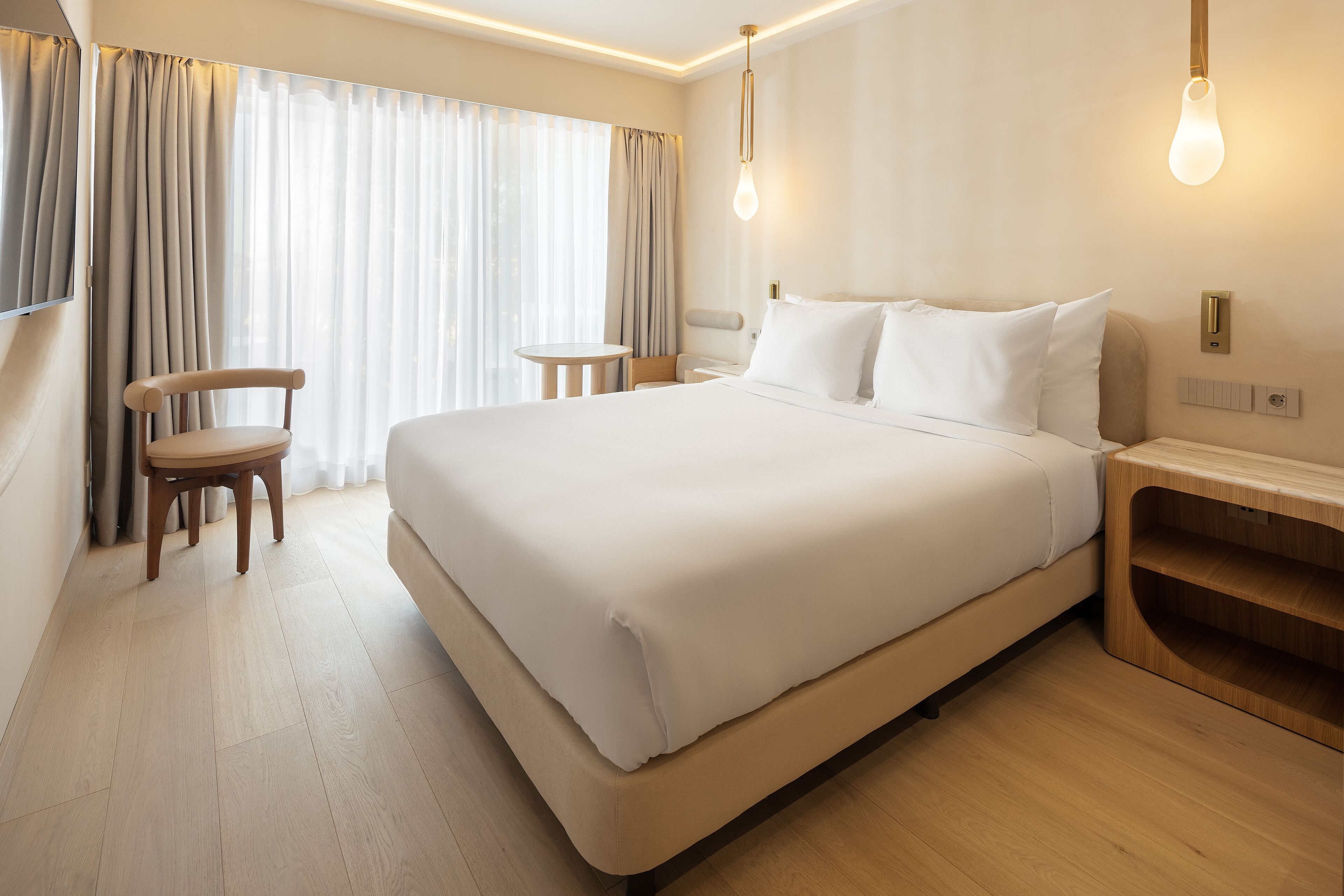 Quarto de hotel com uma cama grande, uma mesinha, uma cadeira e uma mesinha de cabeceira.