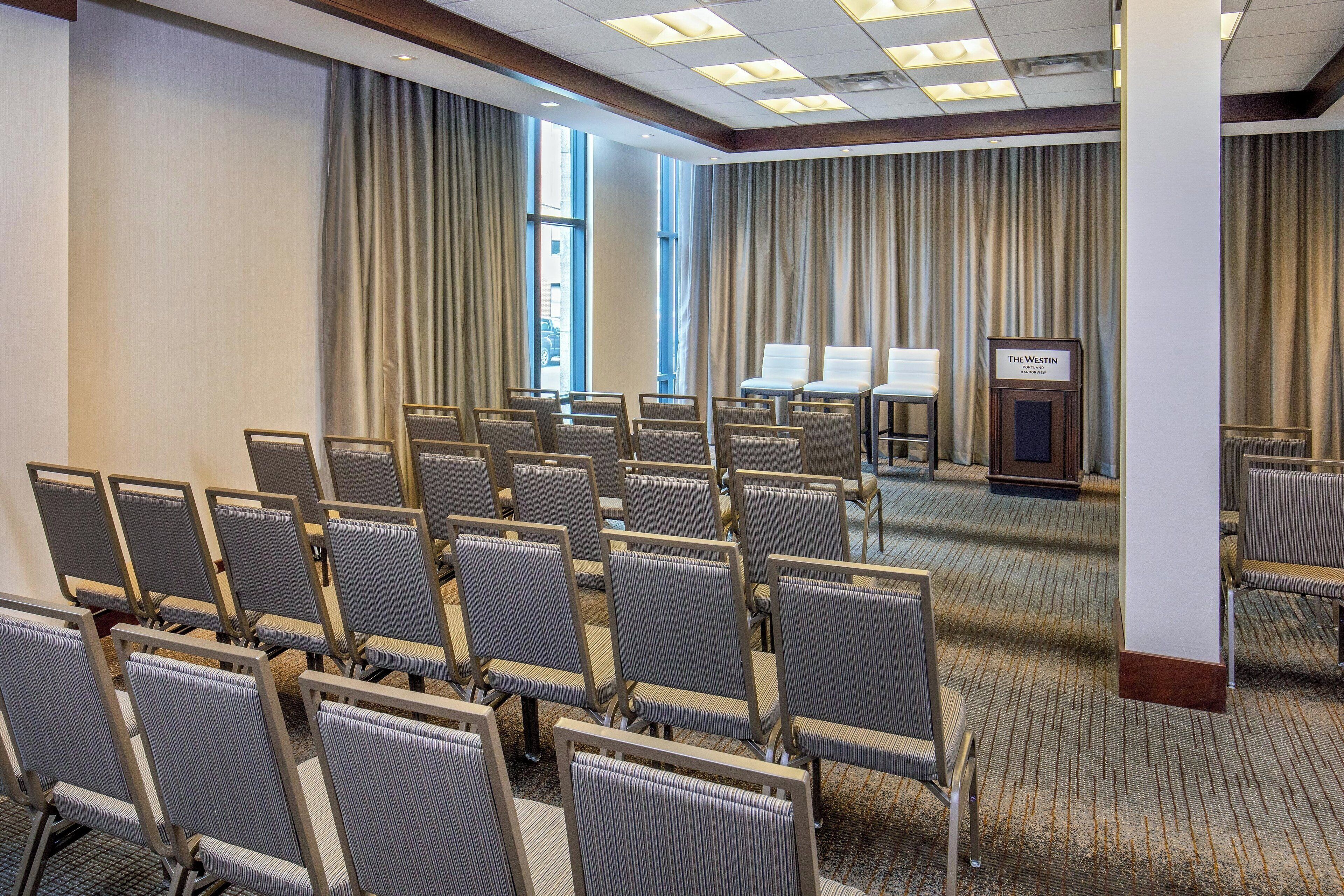 Sala de conferências com fileiras de cadeiras, um púlpito e amplas janelas com cortinas.