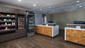 Uma área de recepção de hotel com um balcão de madeira, uma vitrine refrigerada e uma prateleira com lanches.