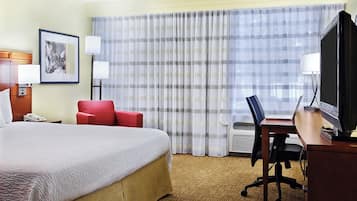 Quarto de hotel com uma cama grande, uma escrivaninha com computador, uma cadeira, uma televisão e uma janela com cortinas.