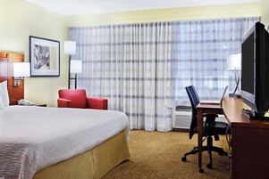 Quarto de hotel com uma cama grande, uma escrivaninha com computador, uma cadeira, uma televisão e uma janela com cortinas.
