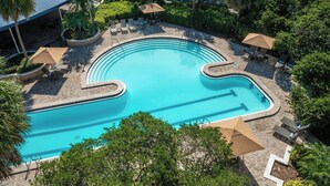 Uma piscina com design singular, rodeada por espreguiçadeiras e guarda-sóis, localizada em uma área ajardinada com árvores e arbustos.