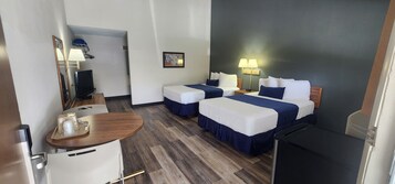 Quarto de hotel com duas camas, uma escrivaninha, uma cadeira, uma mesinha, um espelho e uma luminária na parede.